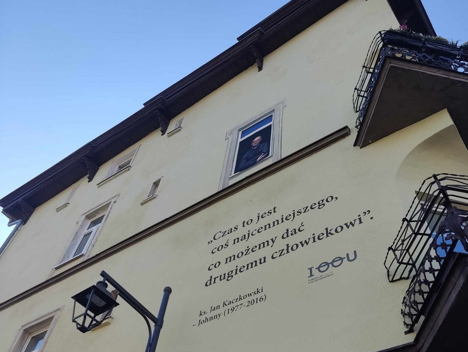 Ksiądz Kaczkowski stoi w oknie i uśmiecha się do przechodniów